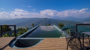 Piscina infinita com deck de madeira e guarda-corpos de metal, com vista para uma paisagem montanhosa.