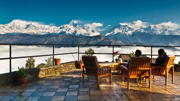 Um terraço com cadeiras e mesa de madeira, com vista para montanhas nevadas e um vale coberto de nuvens.