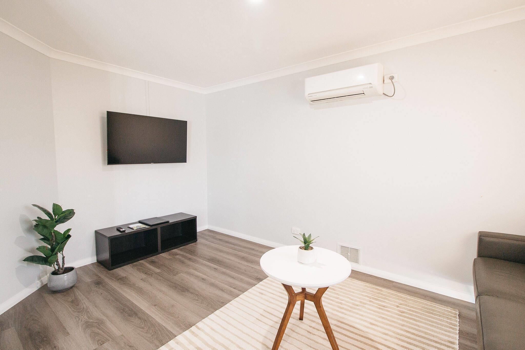 Uma sala de estar moderna com uma televisão de tela plana na parede, um móvel de entretenimento preto, uma mesa redonda branca com uma planta em vaso, um sofá cinza e um tapete estampado no chão.