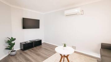 Uma sala de estar moderna com uma televisão de tela plana na parede, um móvel de entretenimento preto, uma mesa redonda branca com uma planta em vaso, um sofá cinza e um tapete estampado no chão.