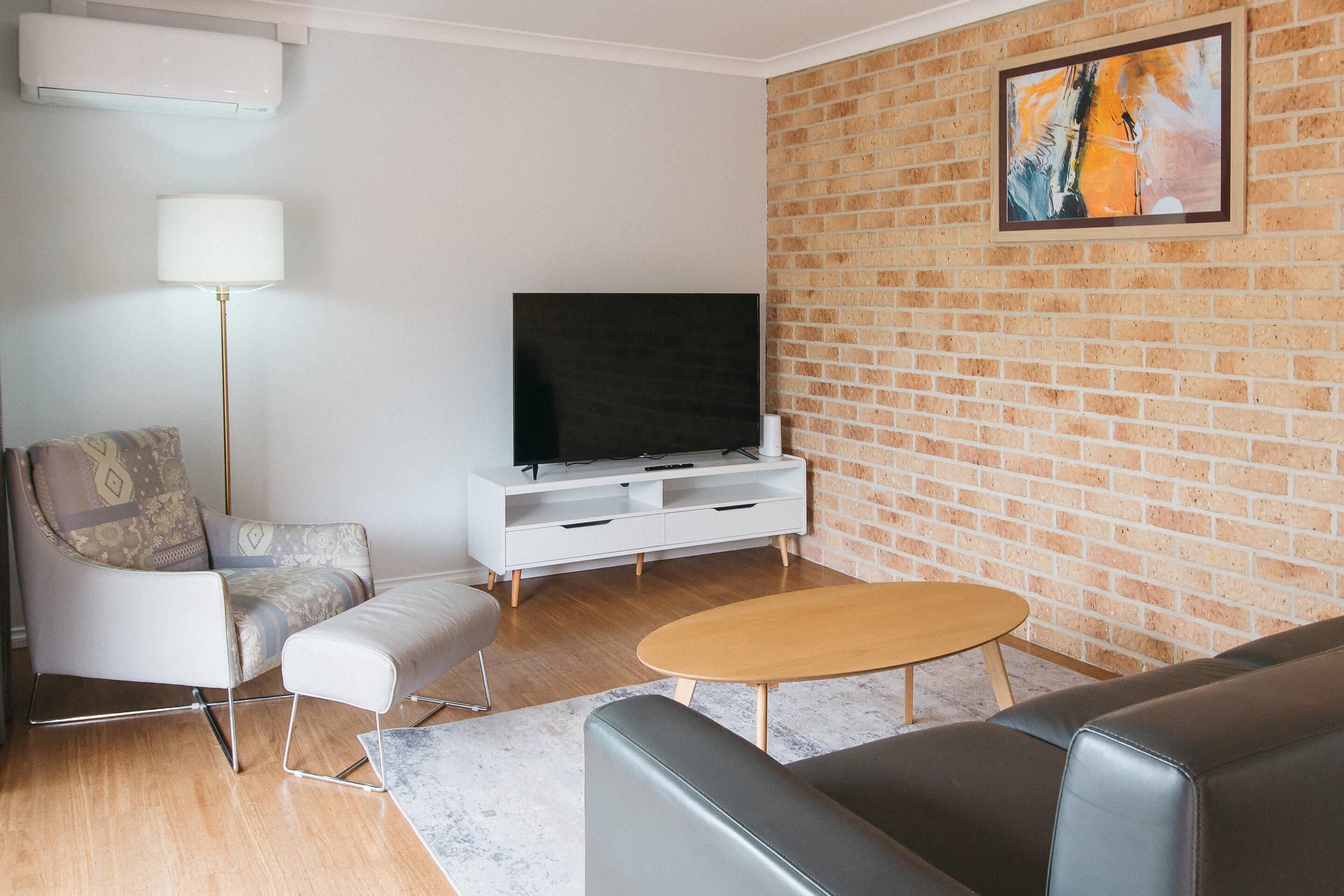 Uma sala de estar moderna com uma televisão de tela plana sobre um móvel branco, um sofá cinza, uma mesa de centro redonda e uma poltrona estampada.