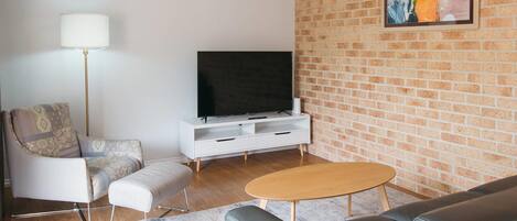 Uma sala de estar moderna com uma televisão de tela plana sobre um móvel branco, um sofá cinza, uma mesa de centro redonda e uma poltrona estampada.