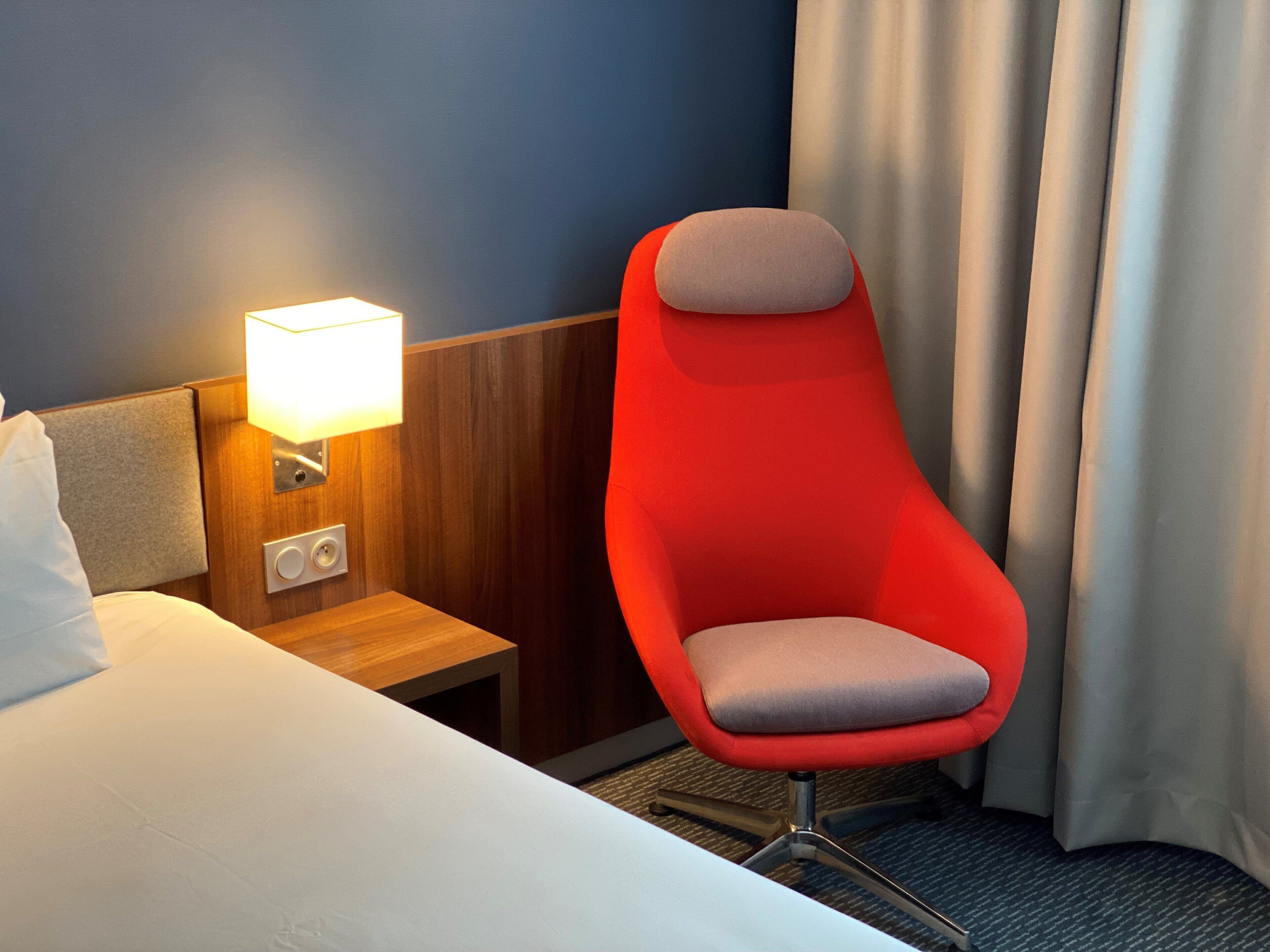 Quarto de hotel com uma cama, uma cadeira vermelha, uma mesa de cabeceira com abajur e uma janela com cortinas.