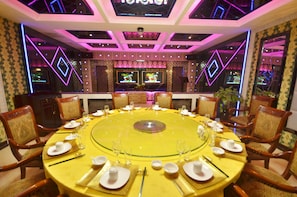 Uma mesa de jantar redonda posta para uma refeição, com cadeiras ao redor, num quarto com papel de parede estampado e iluminação neon no teto.