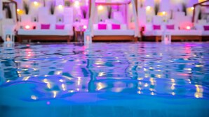 Piscina com reflexo de luzes e uma série de quartos com paredes brancas e detalhes em rosa.