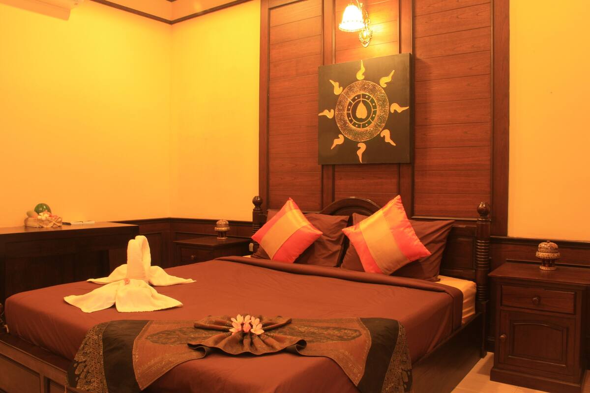 Quarto de hotel com cabeceira de madeira, cama com colcha marrom e duas almofadas, mesa de cabeceira com abajur e uma obra de arte pendurada na parede.
