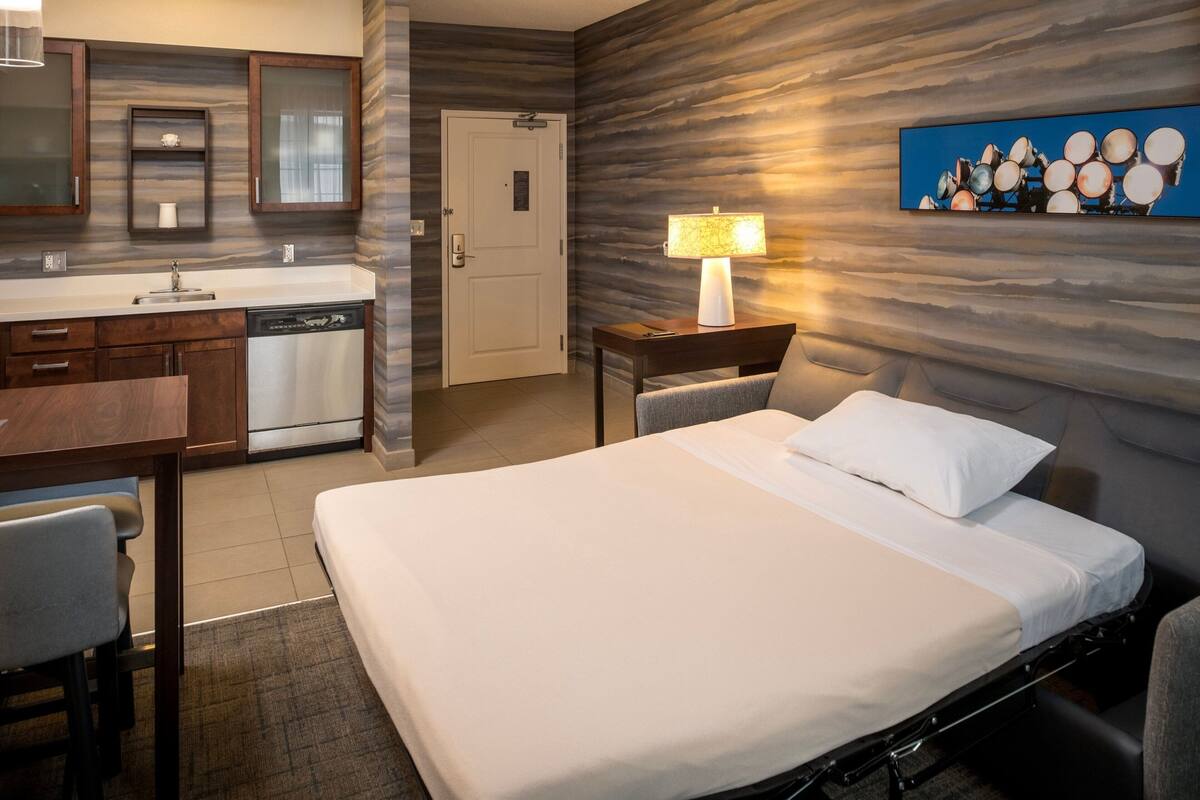Quarto de hotel com cama, escrivaninha e cadeira, mesinha de centro com luminária, banheiro com pia e vaso sanitário, e uma porta.