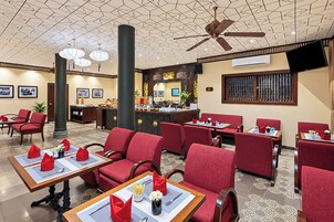 Um espaço de refeições de hotel, com cadeiras estofadas em vermelho, mesas postas com guardanapos e talheres vermelhos, uma área de bar com balcão e banquetas, e um ventilador de teto.
