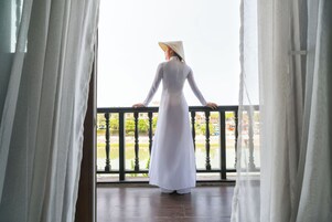 Uma pessoa de vestido branco está em uma varanda com vista para um parque e edifícios.