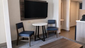 Quarto de hotel com televisão de tela plana na parede, duas cadeiras, uma mesinha redonda e uma mesa de centro de madeira.
