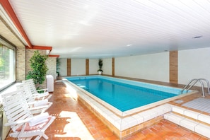 Uma piscina interior com paredes azulejadas e chão de madeira, rodeada por espreguiçadeiras e um vaso de planta.