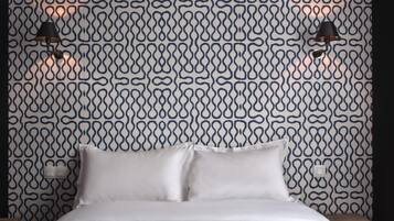 Uma cama com roupa de cama branca, parede estampada e duas luminárias fixadas na parede.