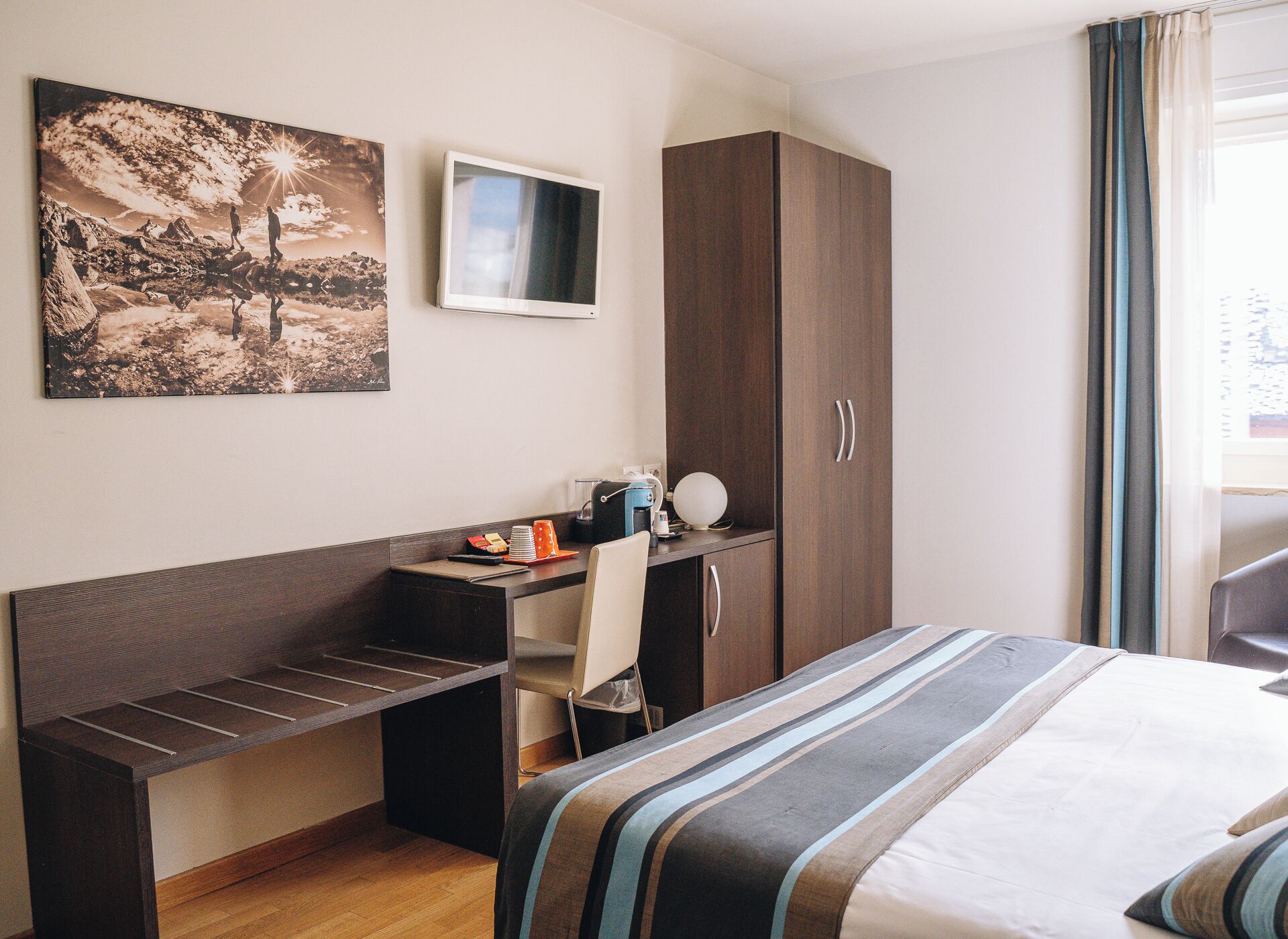 Quarto de hotel com cama, escrivaninha, cadeira, guarda-roupa, televisão de tela plana e um quadro na parede.