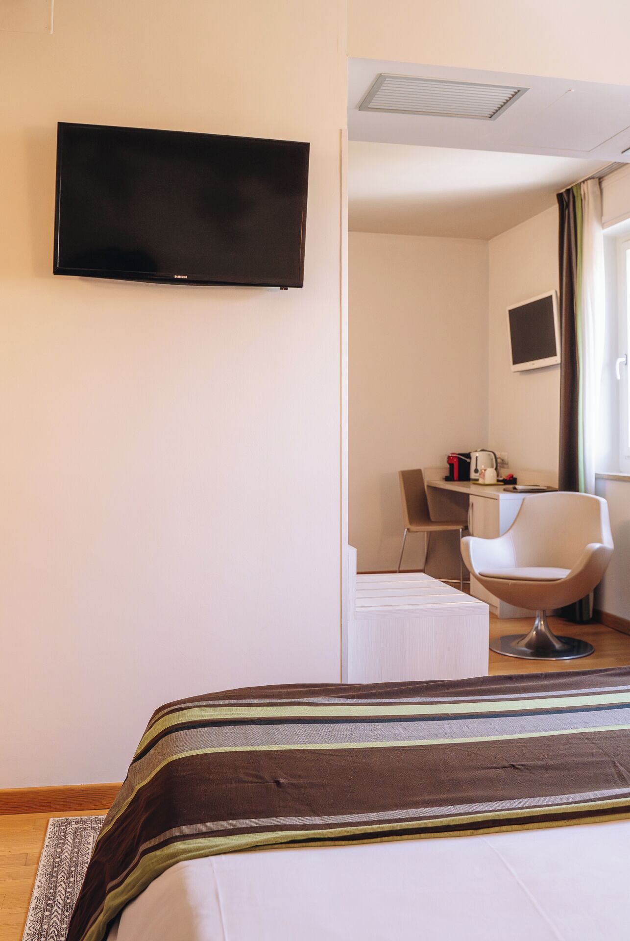 Quarto de hotel com cama, televisão na parede, escrivaninha e cadeira, e janela com cortinas.