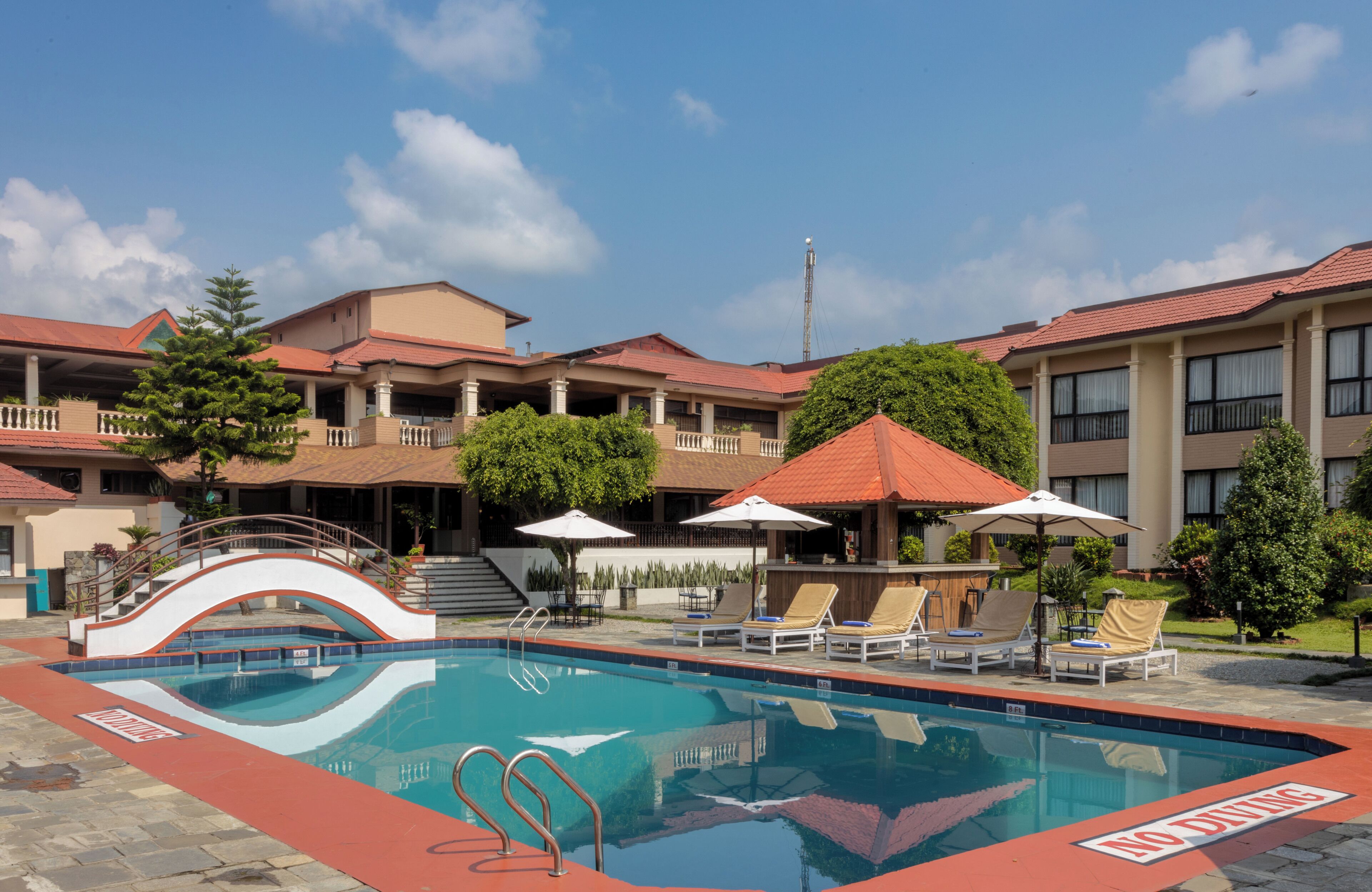 Piscina com espreguiçadeiras e guarda-sóis, cercada por edifícios com telhados vermelhos e céu claro.