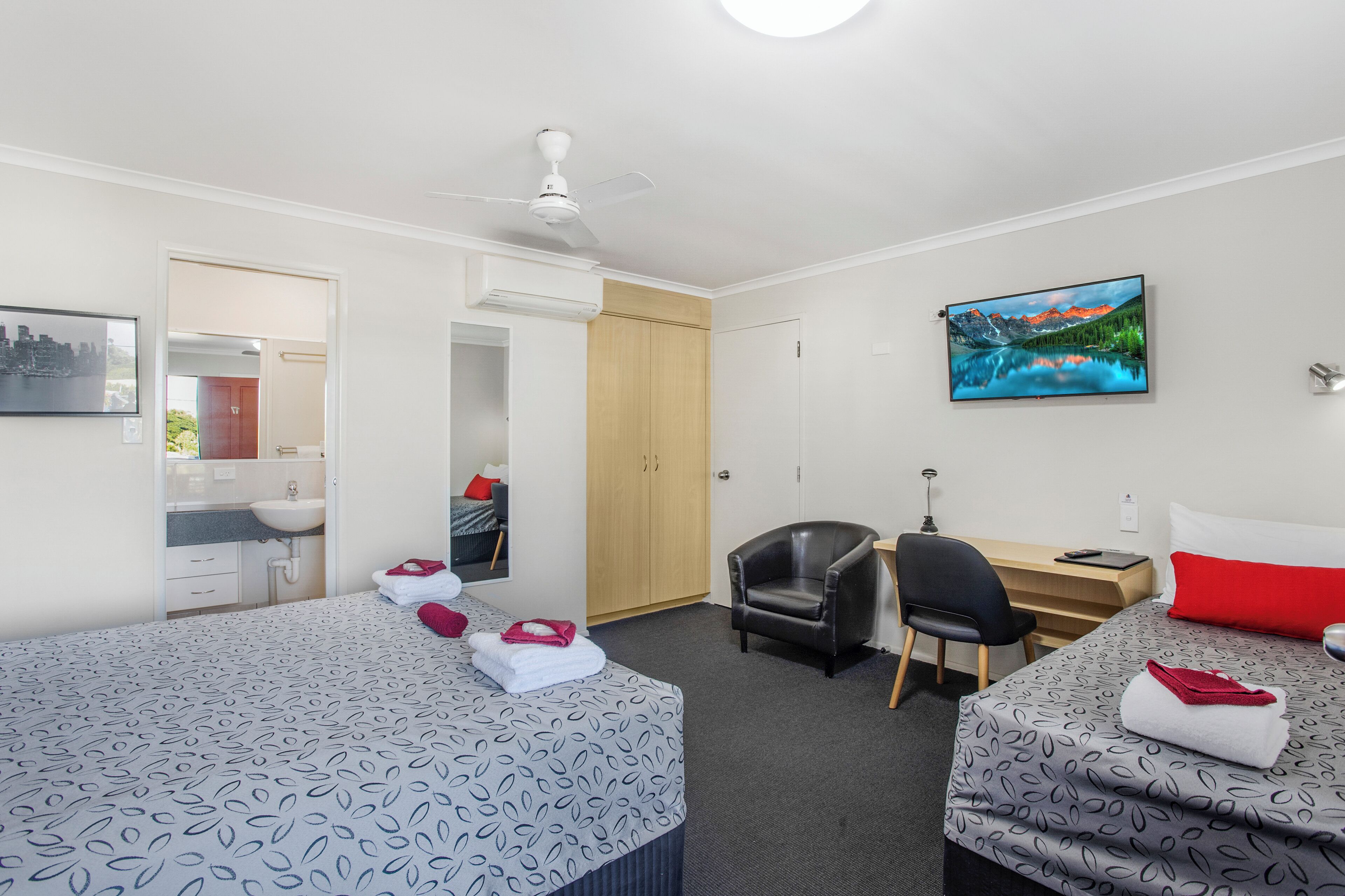 Quarto de hotel com duas camas, uma escrivaninha, uma cadeira, um ventilador de teto e um quadro na parede.