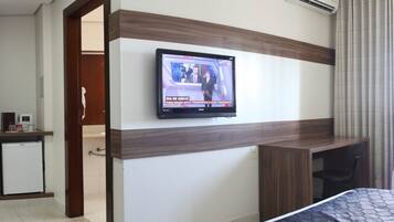 Quarto de hotel com televisão na parede, cama com colcha estampada, pequena escrivaninha com cadeira e guarda-roupa.