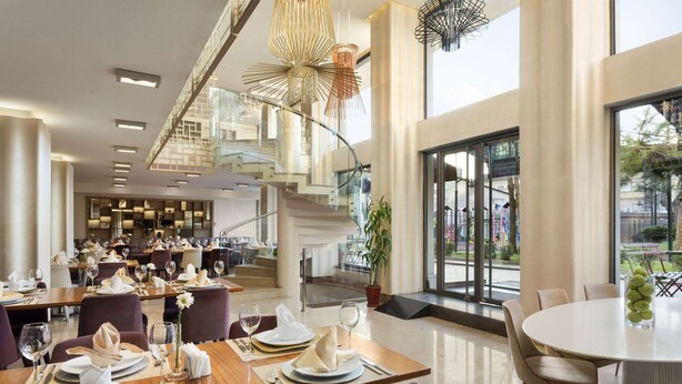 Uma sala de jantar moderna com várias mesas preparadas para hóspedes, uma escadaria que leva a um andar superior e grandes janelas com vista para o exterior.