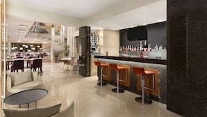 Um hall de hotel moderno com área de bar, disposição de assentos e escadaria que leva aos andares superiores.