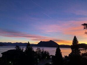 Vista do pôr do sol sobre um lago, com montanhas ao fundo e árvores em primeiro plano.