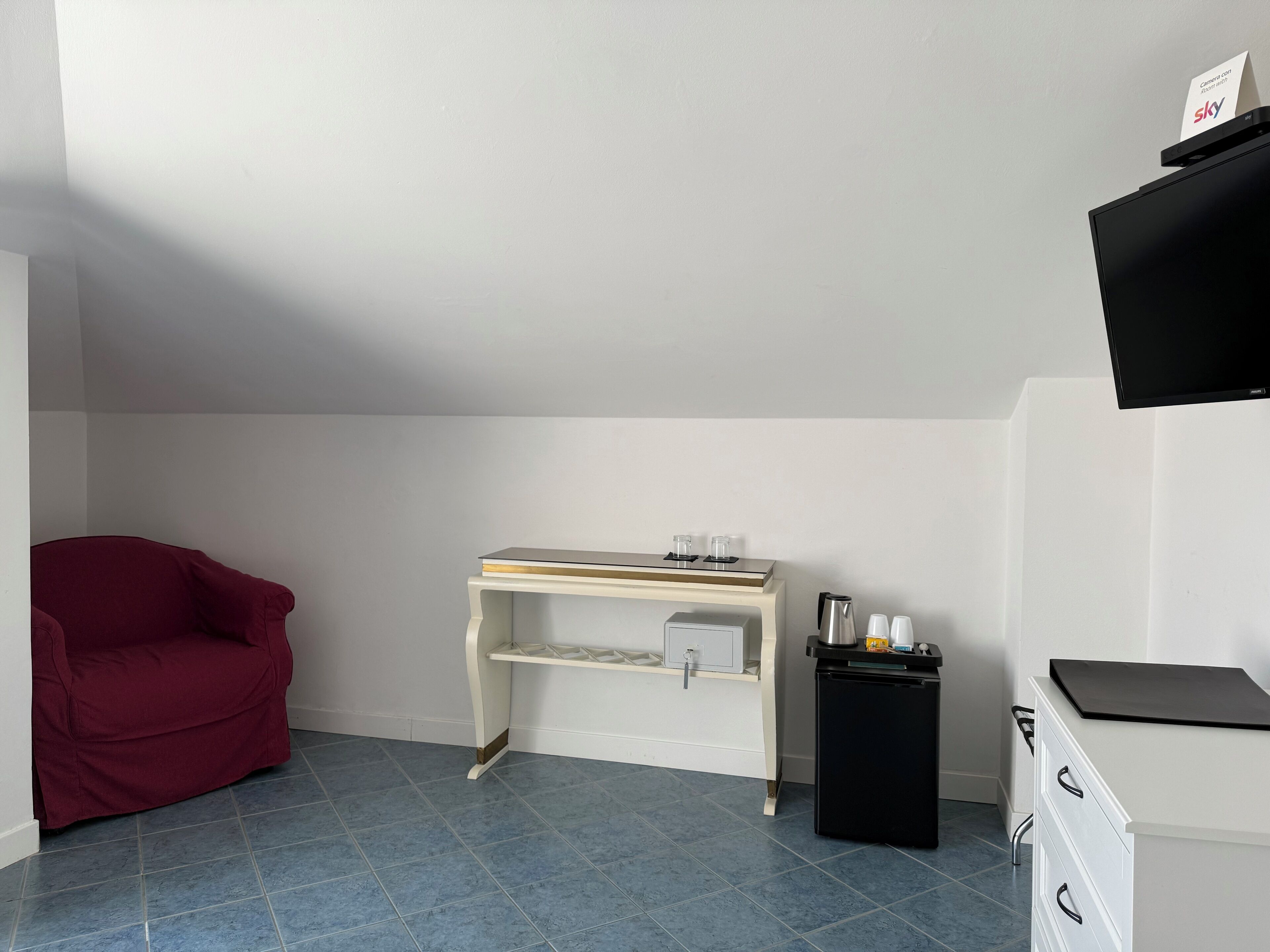 Quarto com escrivaninha, cadeira, pequeno armário preto e televisão presa na parede.