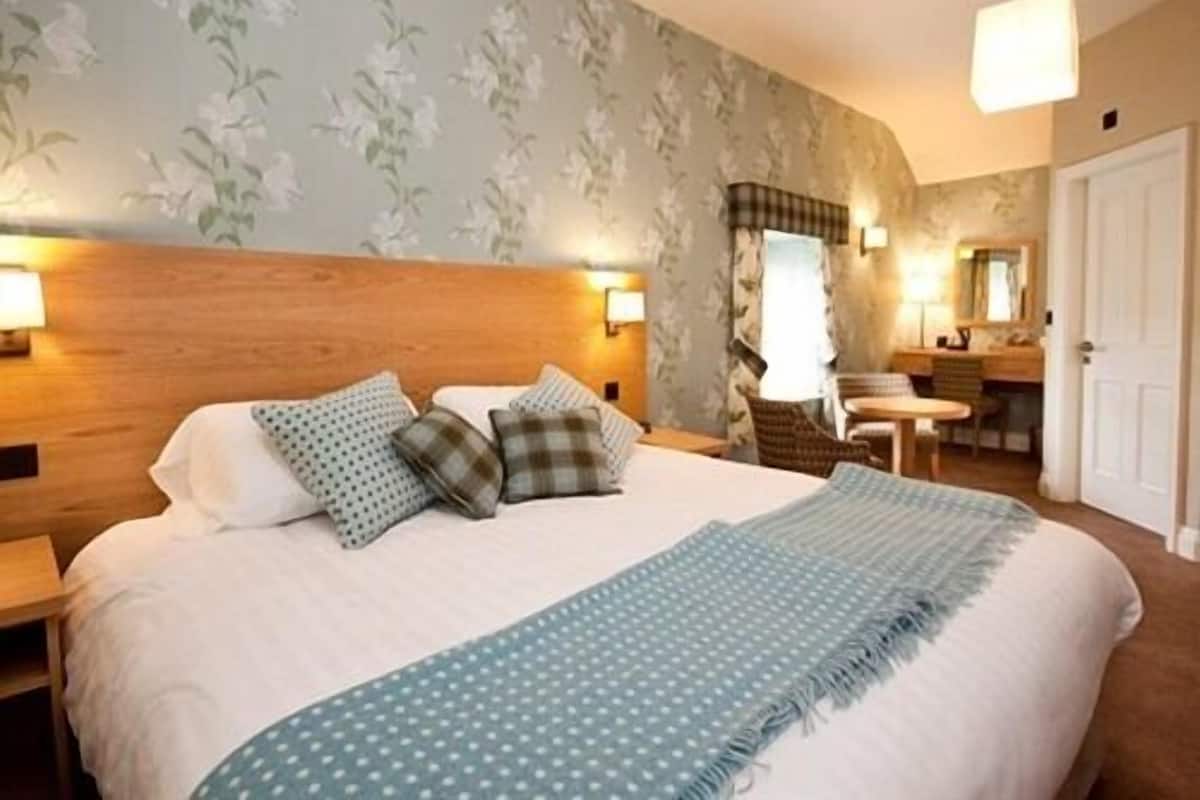 Quarto de hotel com uma cama grande, cabeceira de madeira, escrivaninha com cadeira, banheiro com espelho e uma porta.