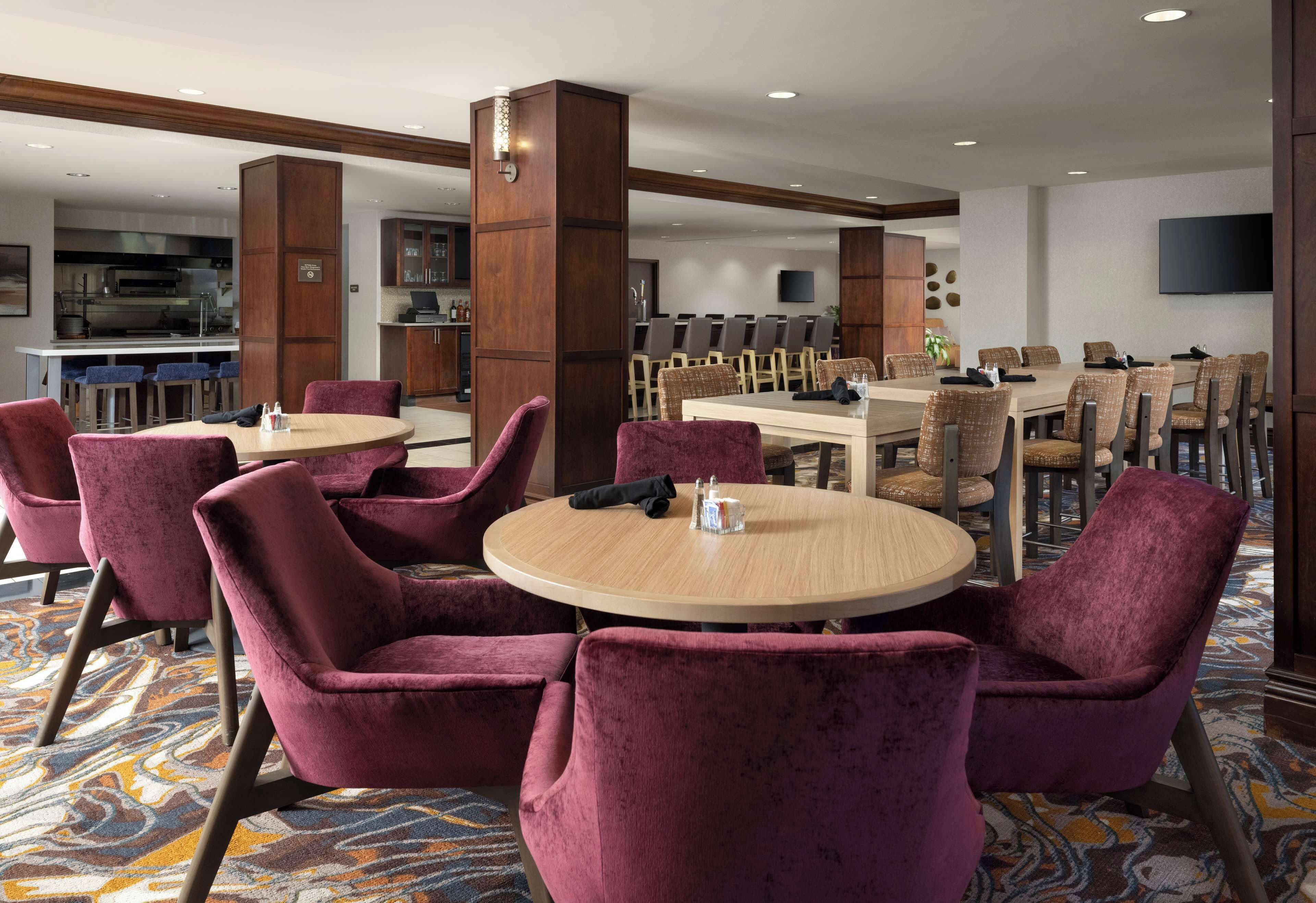 Uma área de refeições de hotel com mesas redondas e cadeiras roxas, um bar e uma televisão fixada na parede.