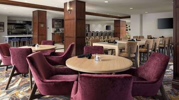 Uma área de refeições de hotel com mesas redondas e cadeiras roxas, um bar e uma televisão fixada na parede.