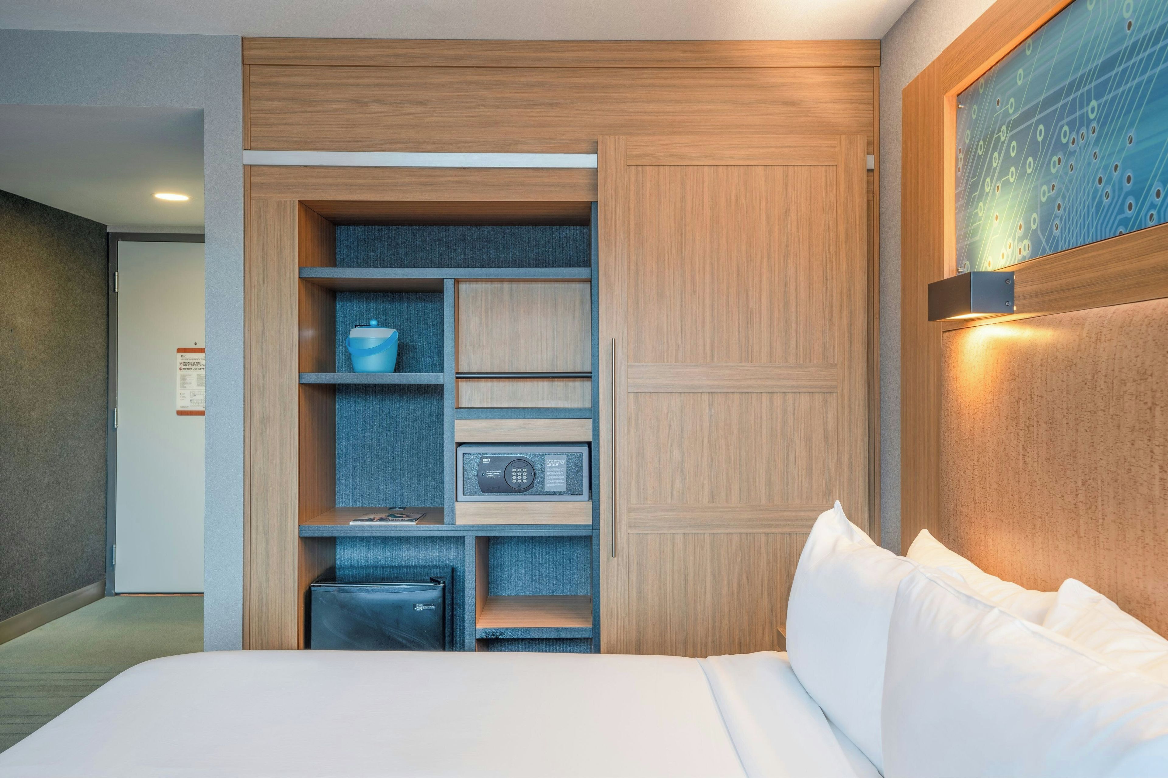Quarto de hotel com cama, roupeiro embutido, secretária com computador e luminária fixada na parede.