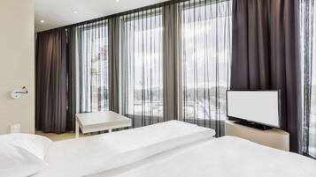 Quarto de hotel moderno com uma cama grande, uma mesinha, uma televisão e janelas amplas com cortinas.