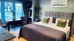 Quarto com uma cama grande, uma escrivaninha e uma cadeira. Há uma janela com persianas, uma planta e um quadro na parede.