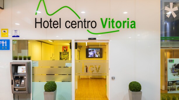 Entrada do Hotel Centro Vitória, com balcão de recepção, uma tela digital e escadaria para o interior.