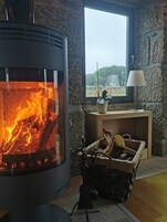 Um quarto aconchegante com parede de pedra, uma lareira grande, uma janela com vista e uma mesinha com abajur.