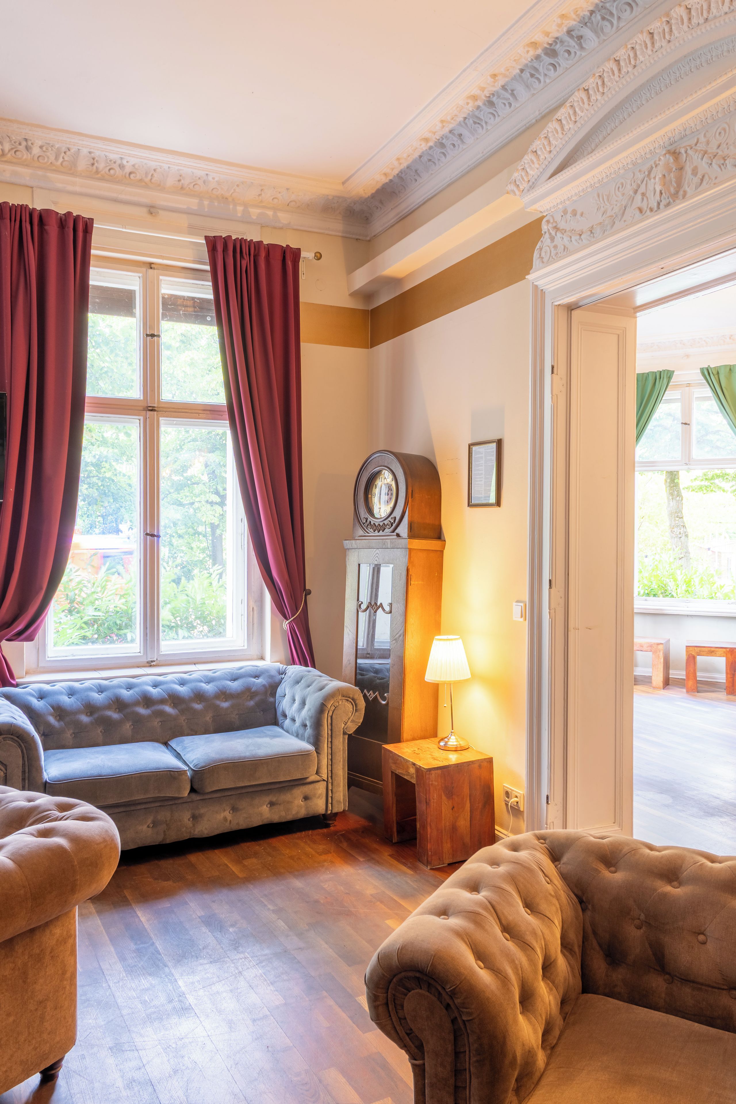 Quarto com sofá azul, poltrona marrom, mesa de cabeceira de madeira com abajur, relógio e janela com cortinas.