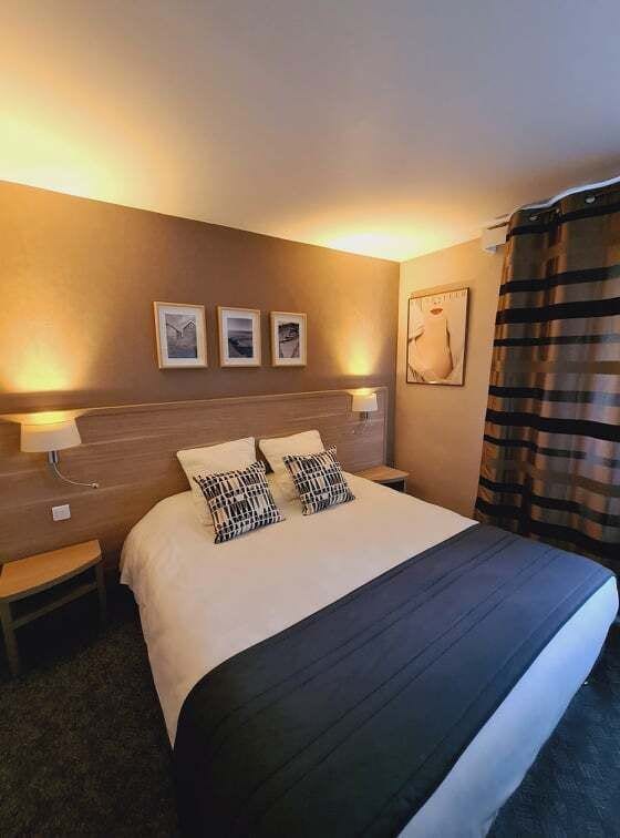 Quarto de hotel com cama, abajures, cabeceira, mesa de cabeceira, um quadro na parede e uma janela com cortina.