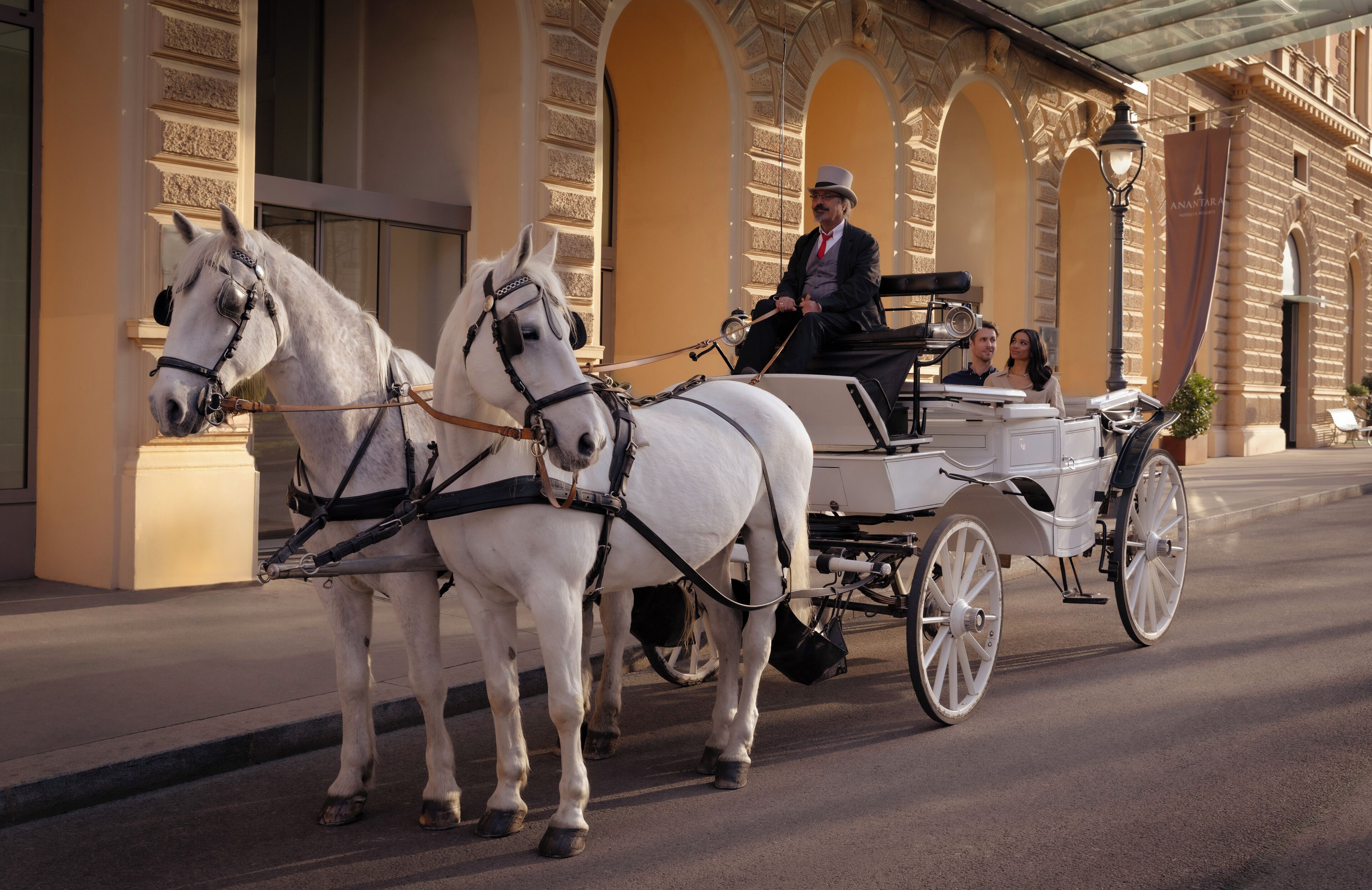 Uma carruagem puxada a cavalo com condutor e passageiros, estacionada em frente a um edifício com janelas em arco e fachada de pedra.