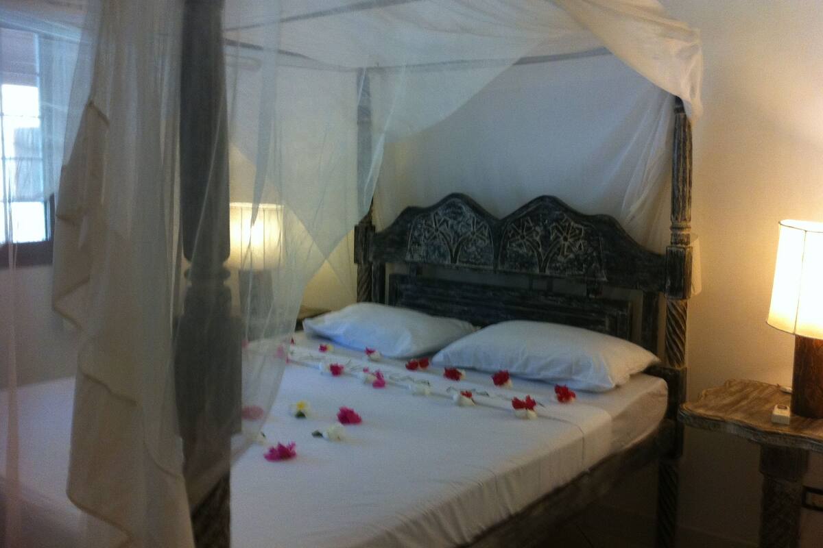 Uma cama com dossel, roupa de cama branca e mesa de cabeceira com abajur.