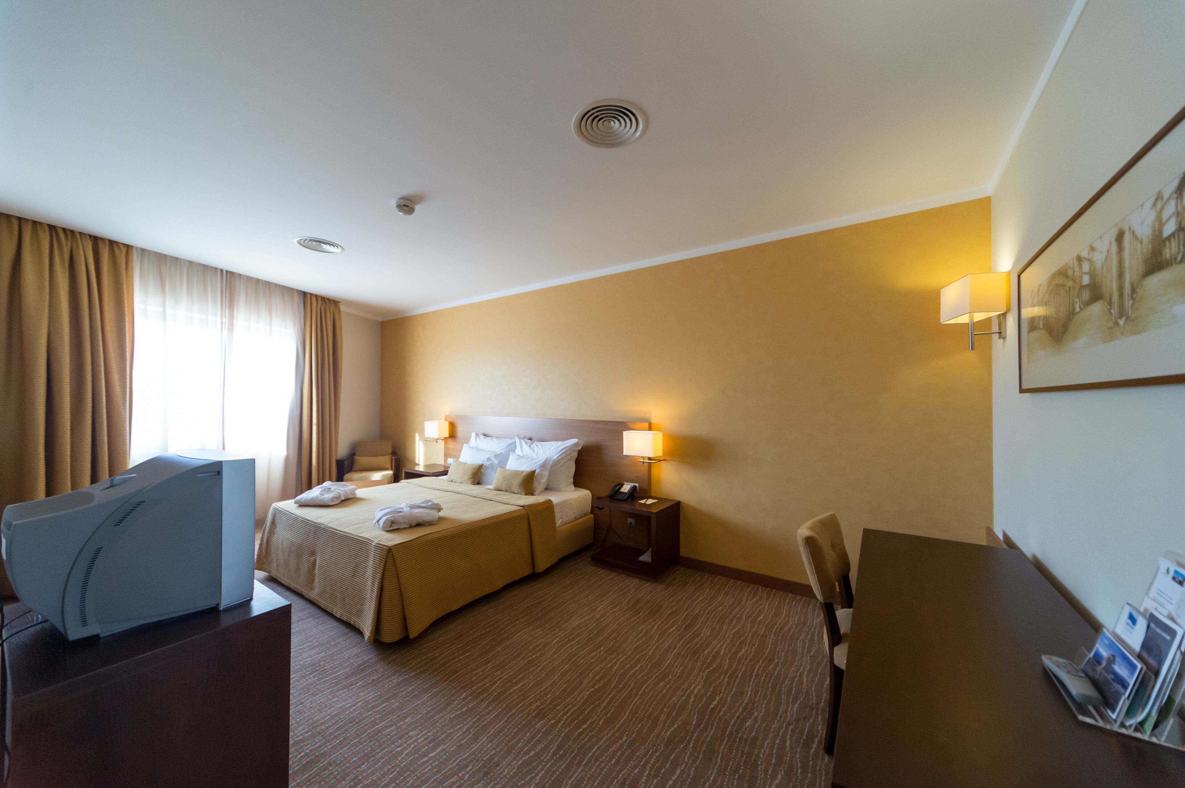 Quarto de hotel com cama, abajures, escrivaninha, cadeira e televisão.