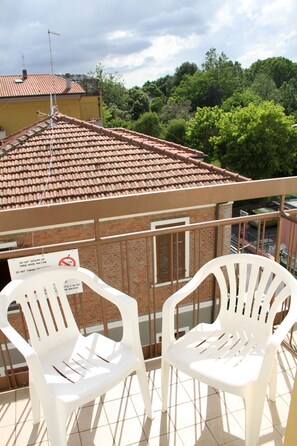 Uma varanda com duas cadeiras de plástico branco, um telhado telhado e um edifício de tijolos ao fundo.