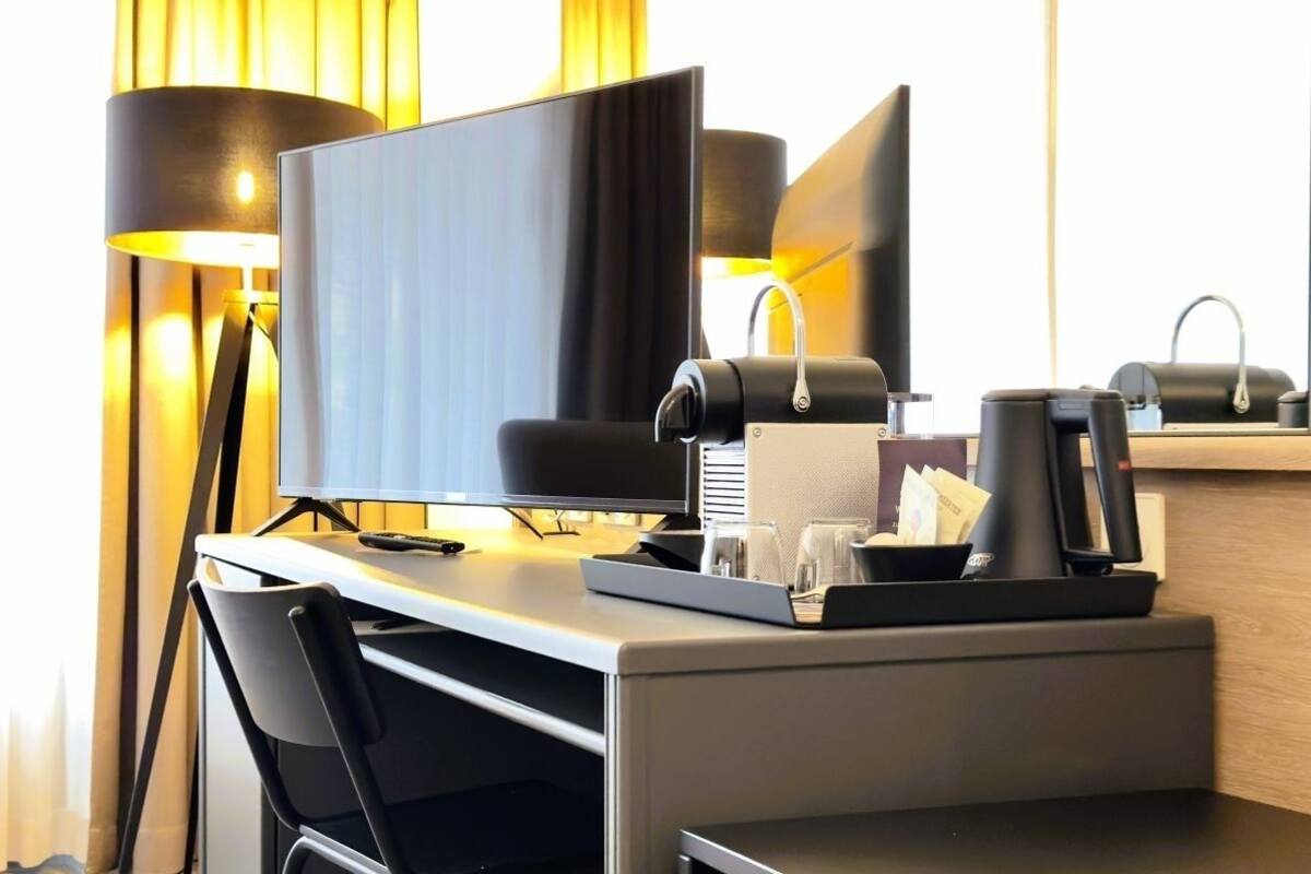Quarto de hotel com escrivaninha, dois monitores, cafeteira e telefone.