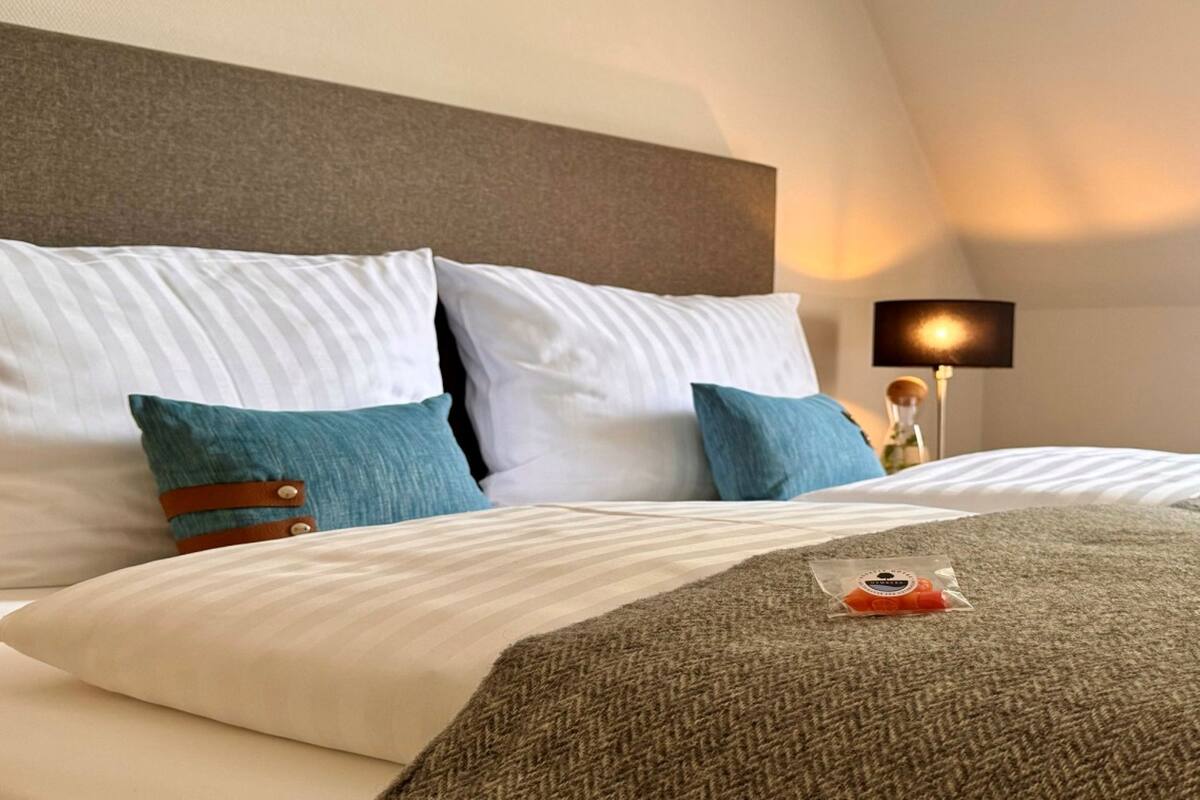Cama bem arrumada com travesseiros brancos e azul-turquesa, cabeceira marrom e abajur no criado-mudo.