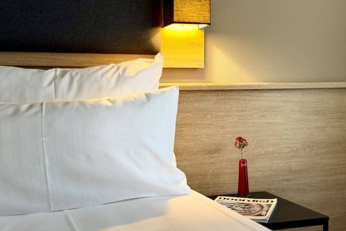 Cama bem arrumada com lençóis brancos, mesa de cabeceira pequena com uma revista e um vaso vermelho com uma flor.