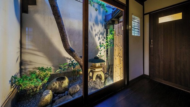Interior japonês tradicional com uma porta de correr que dá para um jardim com arranjo de pedras, um pequeno lampião de pedra e uma cerca de bambu.