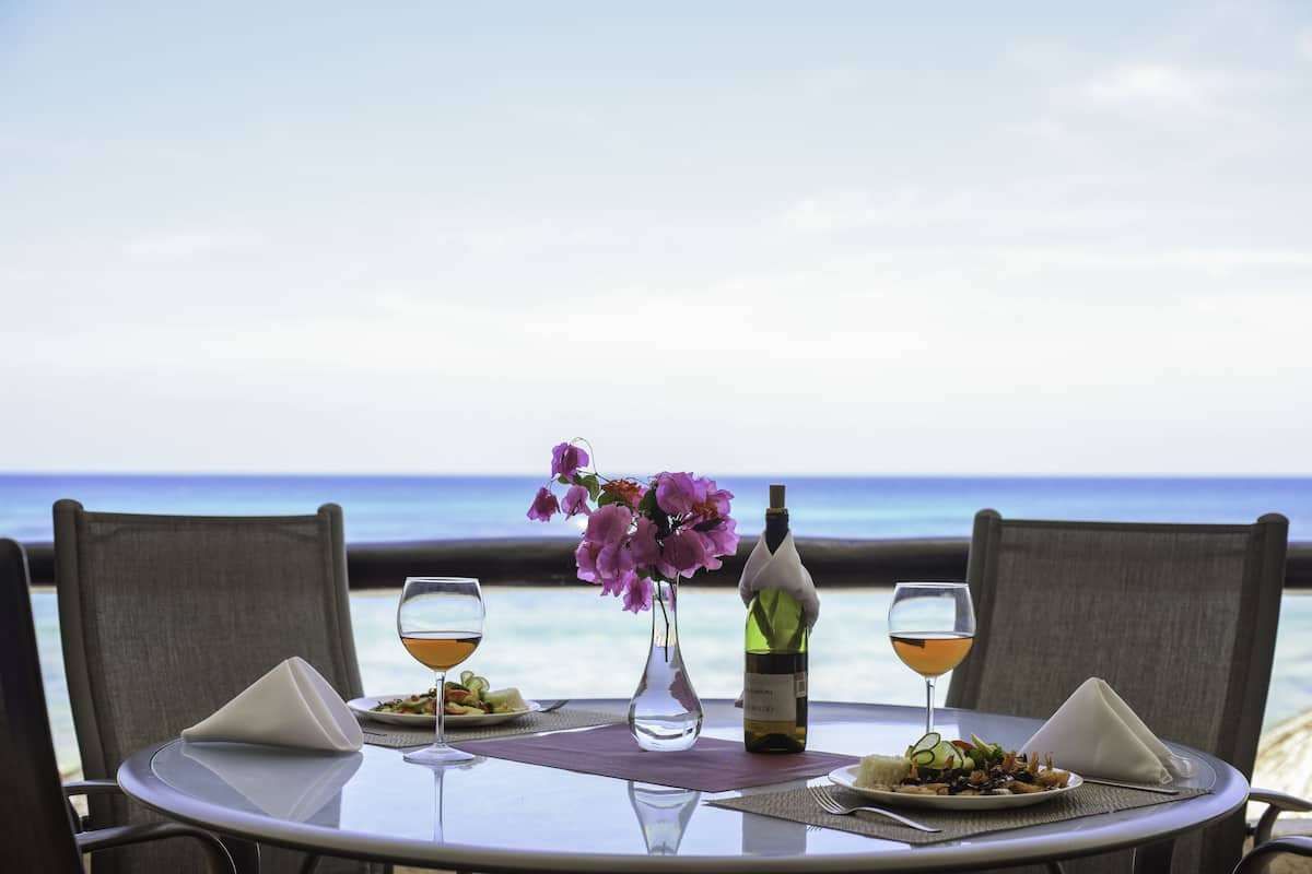Mesa posta para dois com vista para o oceano, uma garrafa de vinho e um vaso de flores.