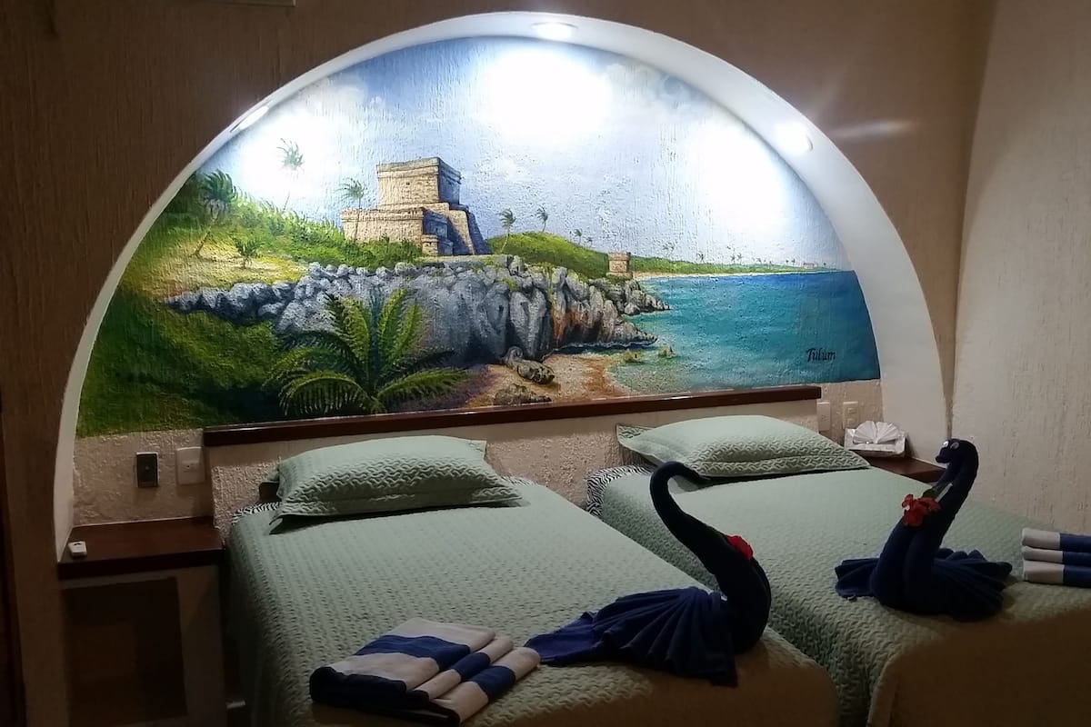 Quarto com duas camas, um mural com paisagem costeira e uma pequena estátua na cama.