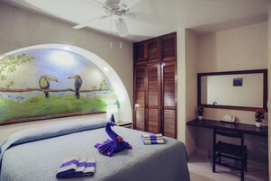 Quarto de hotel com cama, escrivaninha, cadeira, ventilador de teto e uma grande pintura de tucanos na parede.