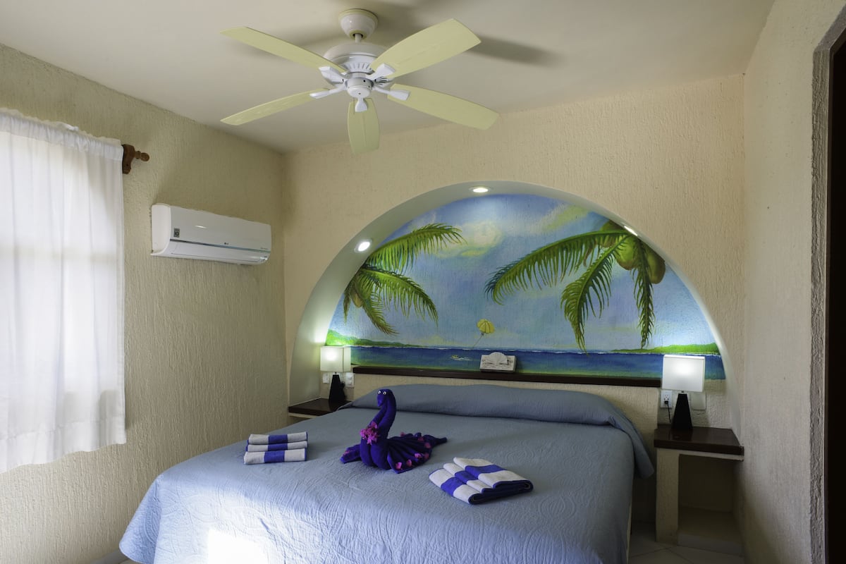 Quarto de hotel com uma cama, um ventilador de teto, um aparelho de ar condicionado, uma pintura de uma cena de praia e uma mesa de cabeceira com uma luminária.