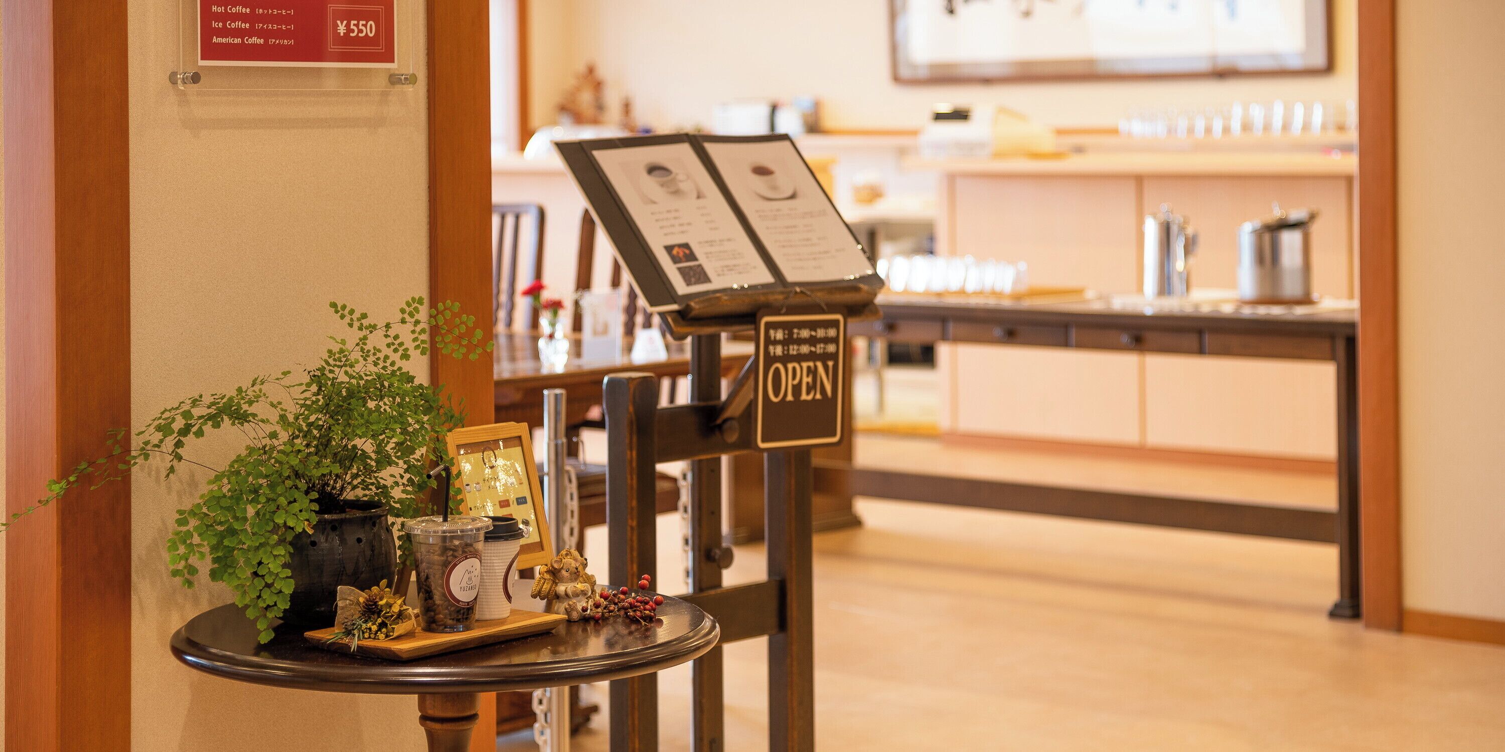 Uma área de recepção com uma cadeira de madeira, uma mesinha com uma planta, um suporte para cardápio e um aviso indicando que o café está aberto.