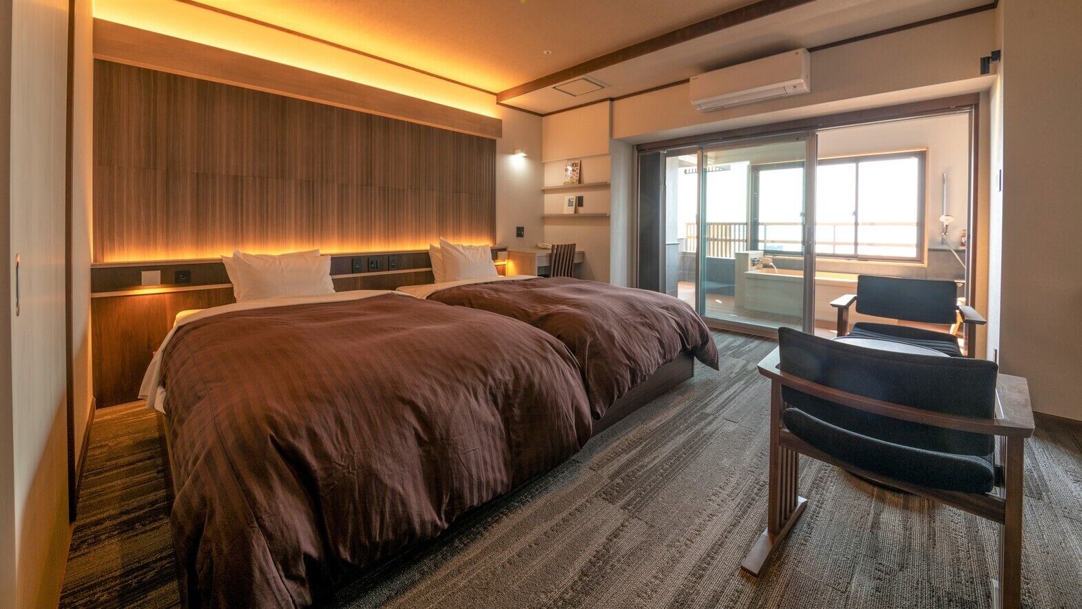 Quarto de hotel com uma cama grande, uma escrivaninha com cadeira, porta de vidro deslizante para uma varanda e uma estante embutida com objetos decorativos.