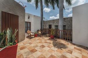 Um pátio com piso xadrez, mesas e cadeiras ao ar livre, plantas em vasos e um edifício de pedra.